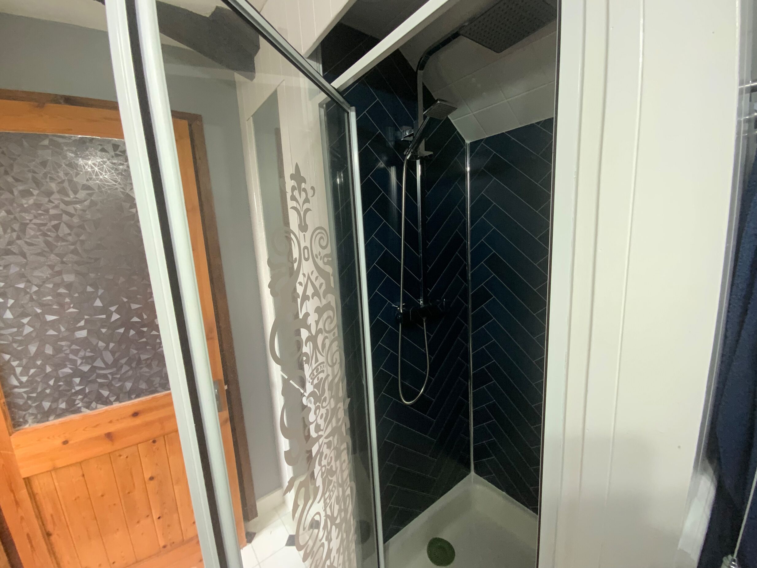 Um chuveiro com box de vidro, parede revestida com azulejos pretos e moldura de porta de madeira.