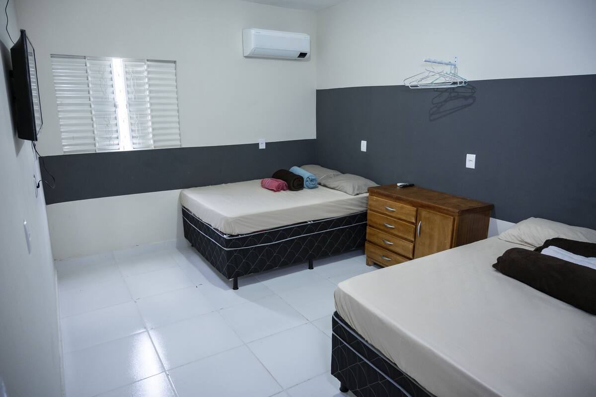 Quarto com duas camas, cómoda de madeira, ar condicionado fixado na parede e janela com persianas.