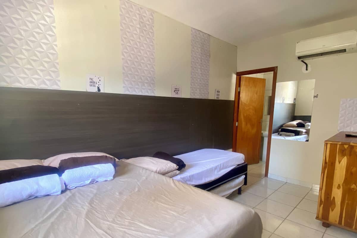 Quarto de hotel com duas camas, cabeceira de madeira, piso de azulejos e um pequeno armário de madeira.
