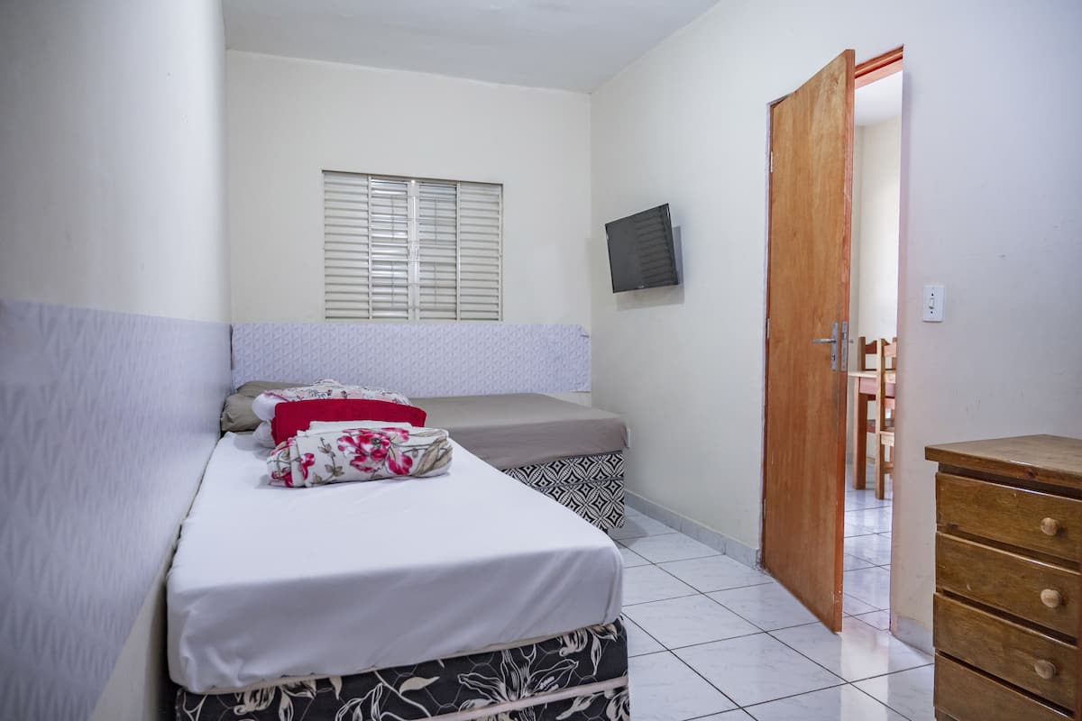 Quarto com cama, mesa de cabeceira, televisão fixada na parede e porta de madeira que dá para outro cômodo.