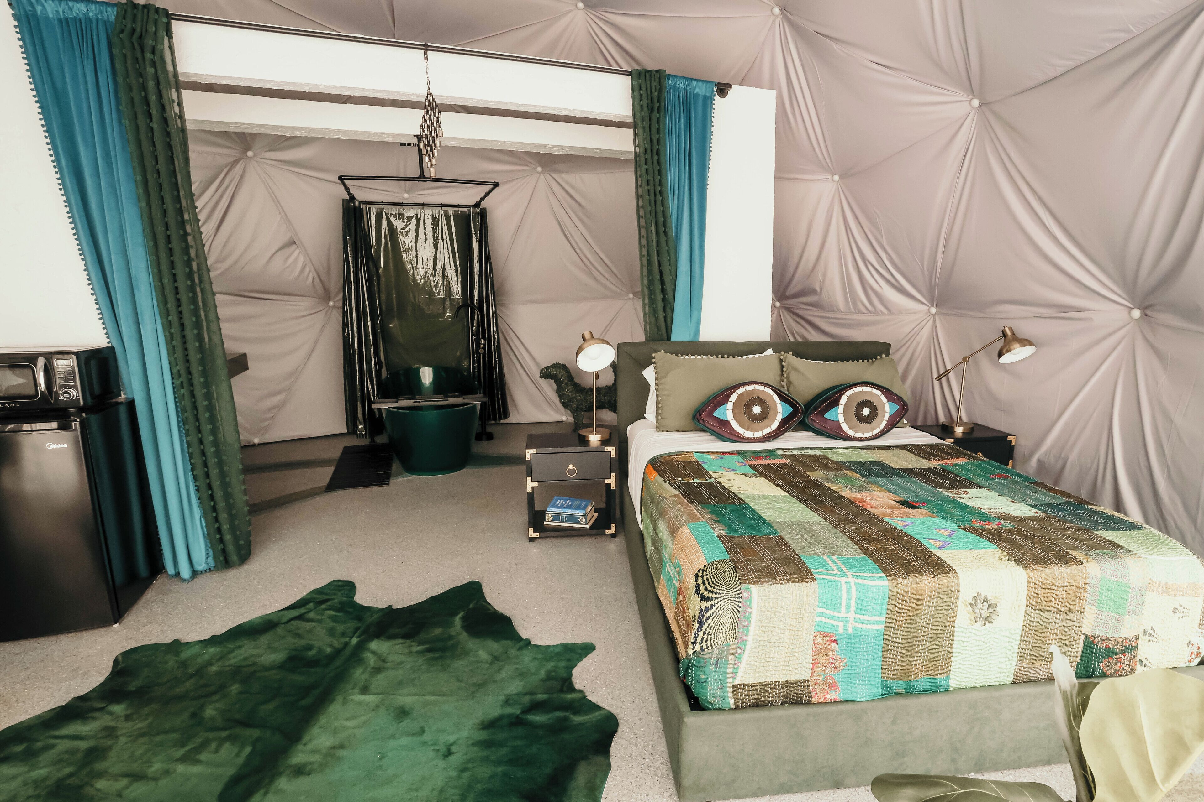 Quarto com cabeceira estofada, edredom acolchoado, tapete verde e pequena mesa de cabeceira com abajur.
