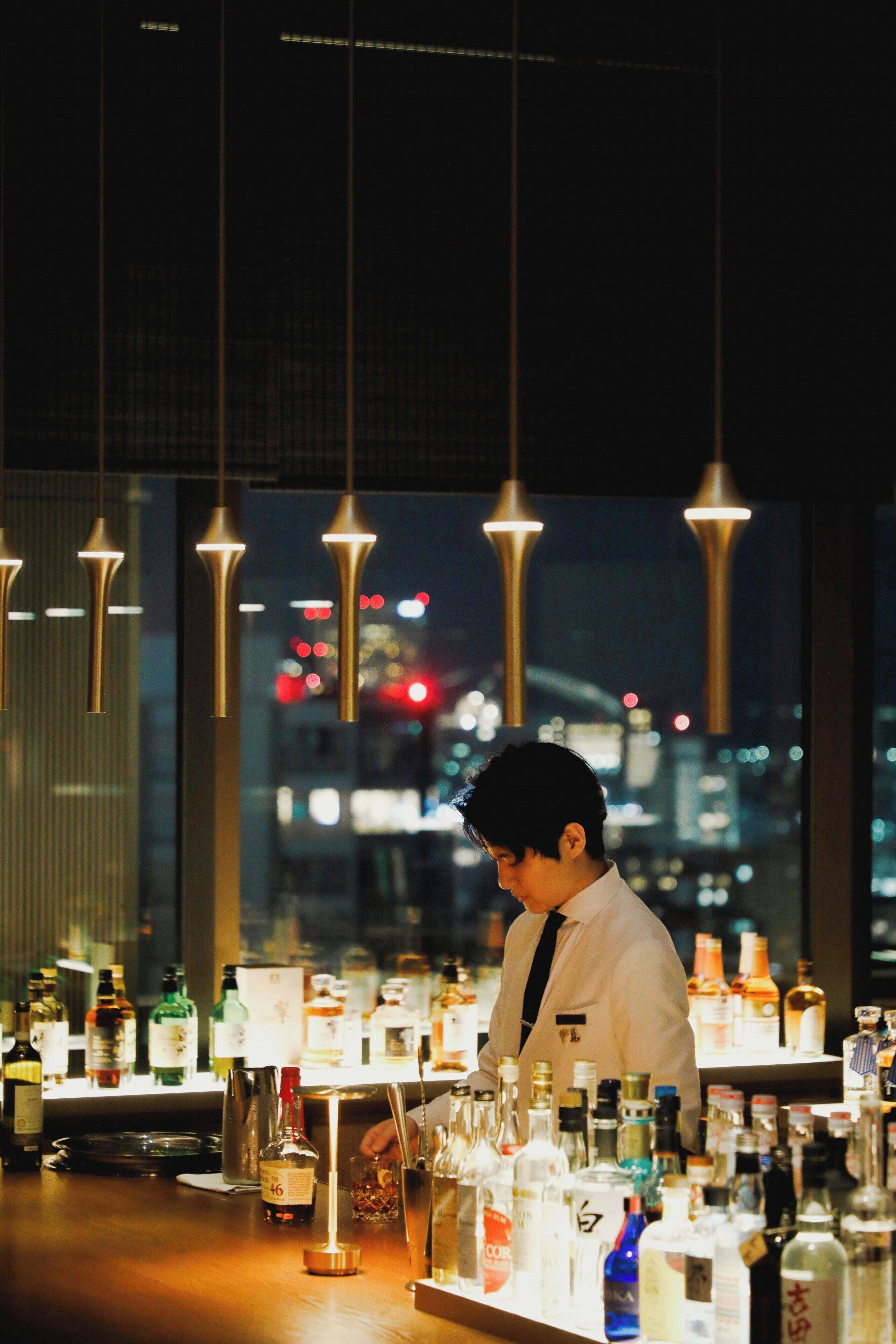 Um bar com vista para a cidade, garrafas de álcool e luminárias penduradas.
