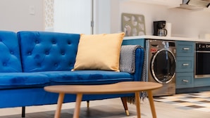 Uma sala de estar moderna com um sofá azul, uma mesa de centro de madeira e uma máquina de lavar ao fundo.