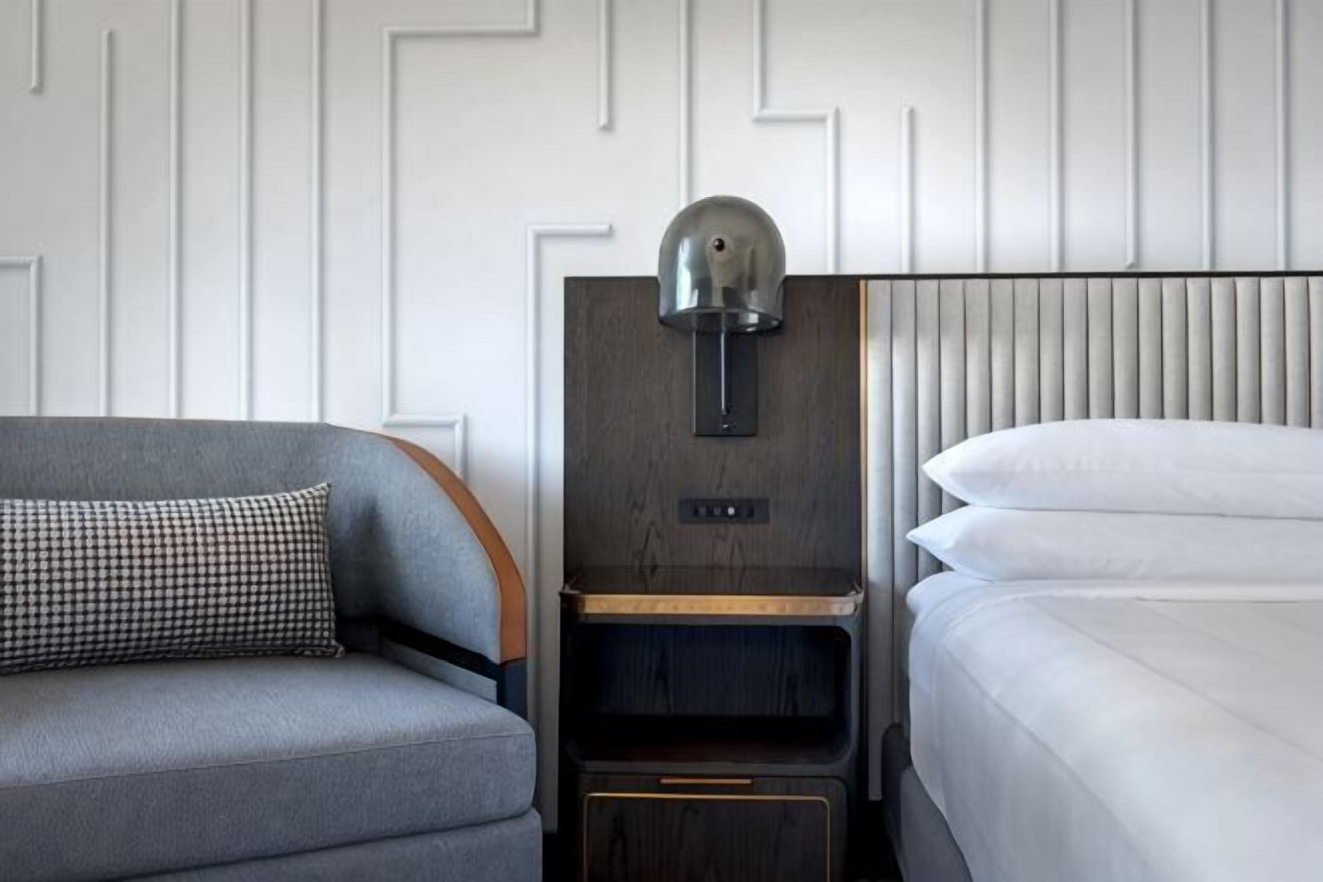 Quarto de hotel moderno com sofá cinza, mesa de cabeceira de madeira com abajur e cama bem arrumada.