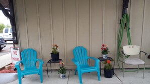Duas cadeiras turquesa, uma cadeira branca e uma mesa pequena com plantas em vasos em frente a um edifício com placa de 'NÃO ESTACIONAR'.