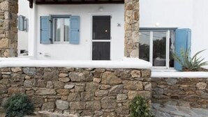 Um edifício moderno branco com paredes de pedra e contraportas azuis, apresentando entrada com piso de pedra e área externa coberta.