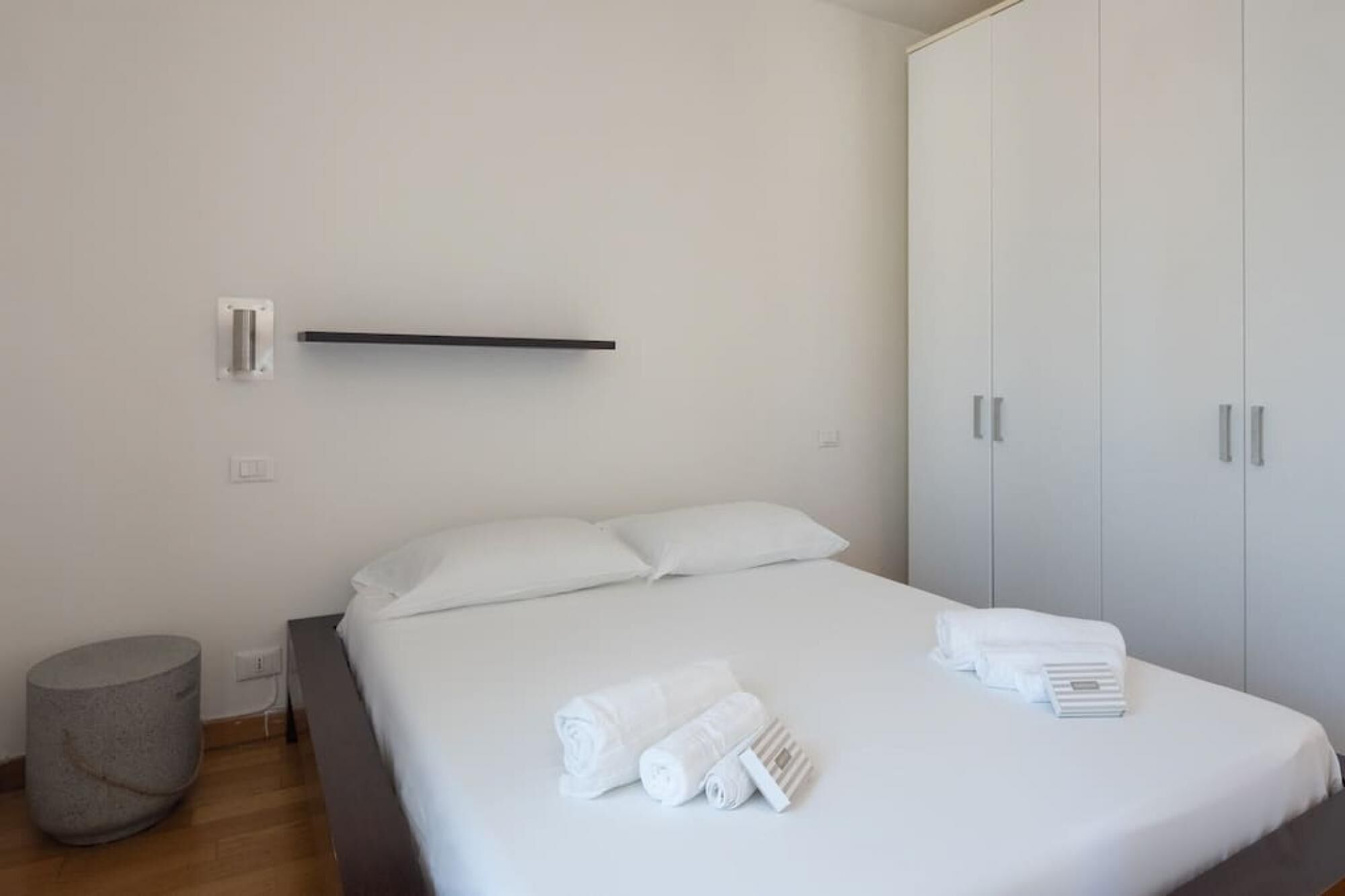 Quarto de hotel minimalista com uma cama, um guarda-roupa e um pequeno banquinho redondo.