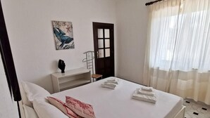 Quarto de hotel com cama, pequena escrivaninha, cadeira, porta e janela com cortinas.