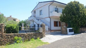 Uma casa de dois andares com um portão de metal, uma entrada em muro de pedra e uma grande árvore em frente.