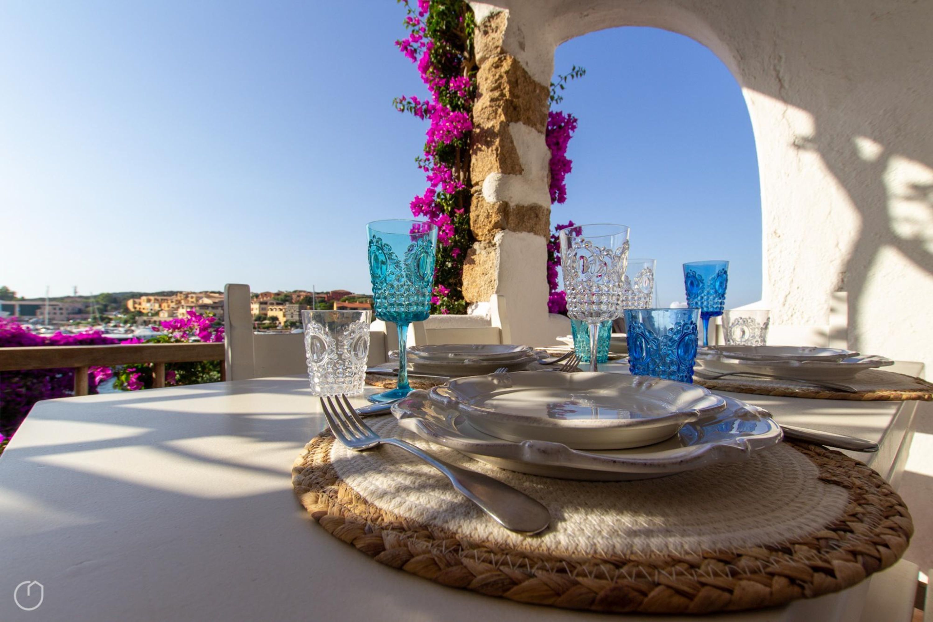 Mesa posta para dois com copos, pratos e talheres azuis, com vista panorâmica para flores cor-de-rosa e edifícios.