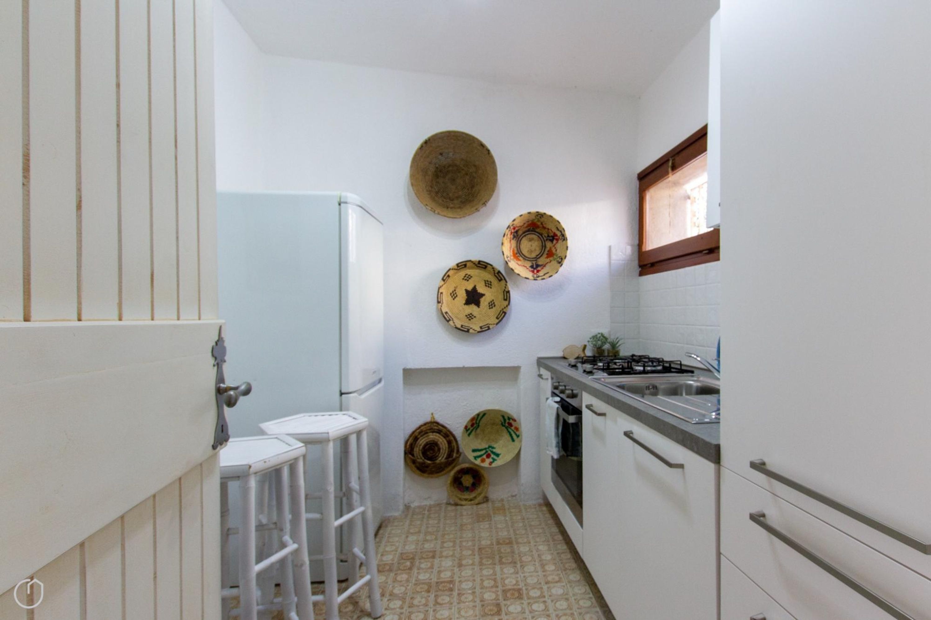 Uma cozinha compacta com armários brancos, fogão a gás, pia e geladeira. Há banquetas e pratos decorativos na parede.