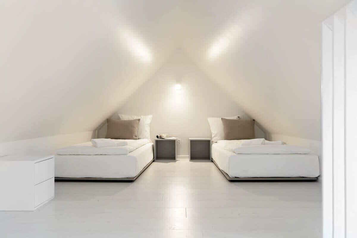Quarto moderno e minimalista com duas camas brancas, uma pequena mesa de cabeceira e um armário branco.
