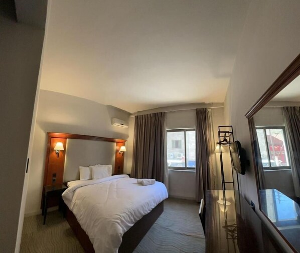 Quarto de hotel com uma cama grande, uma escrivaninha, uma cadeira, uma televisão e uma janela com cortinas.