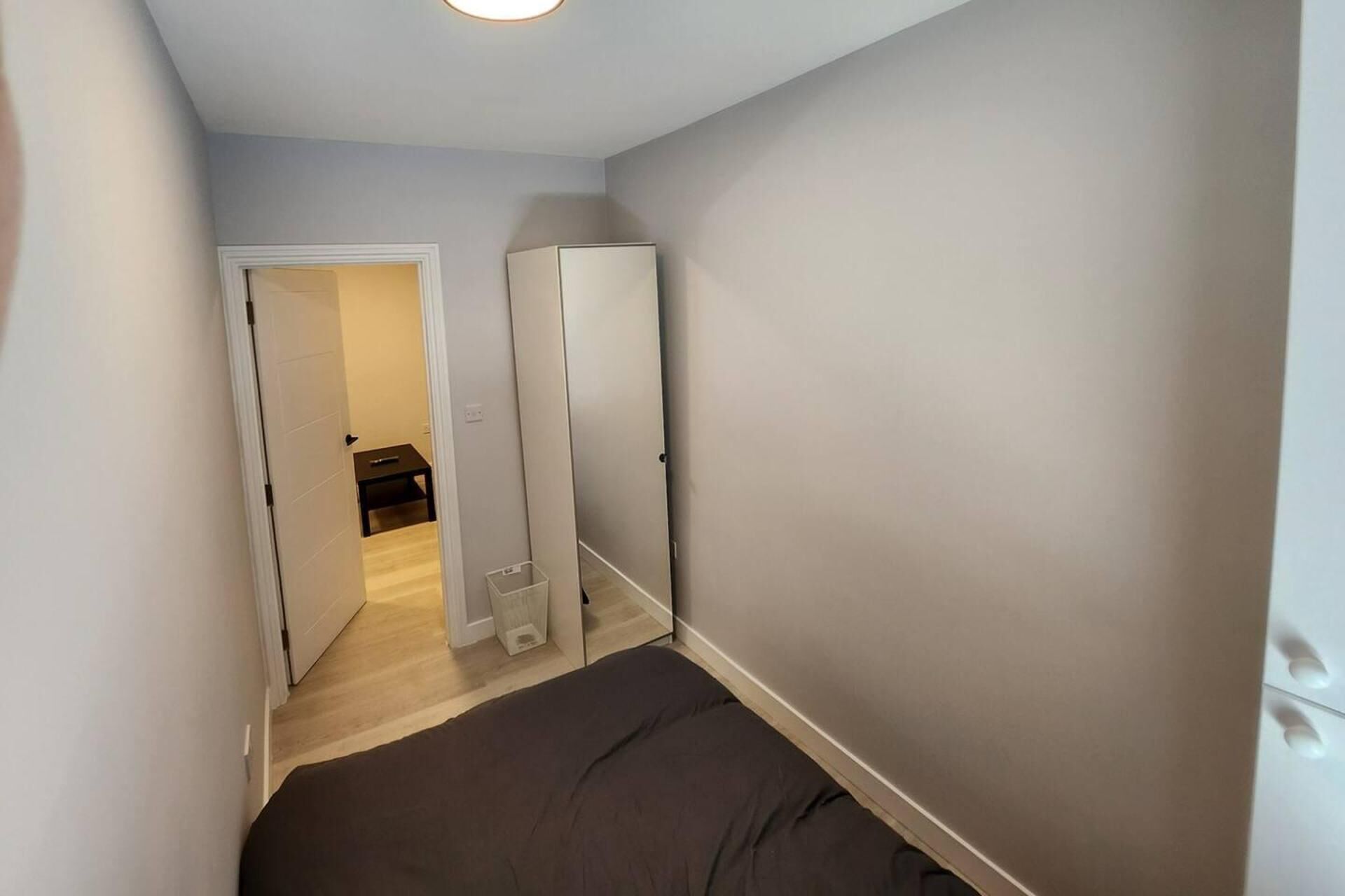 Um corredor estreito com uma cama no fim, uma porta que dá para outro quarto e uma pequena mesa com uma cadeira.