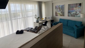 Uma sala de estar moderna com um sofá azul, uma área de jantar com mesa e cadeiras, e uma janela grande com cortinas leves.