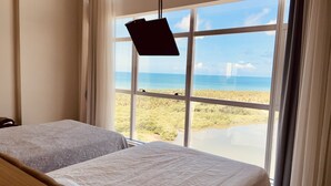 Quarto com duas camas, uma janela ampla com vista para a praia e oceano, e um lustre.