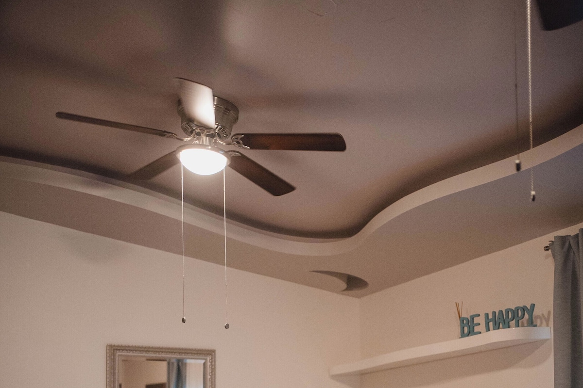 Quarto com cama, mesas de cabeceira, ventilador de teto, espelho e um letreiro na parede.