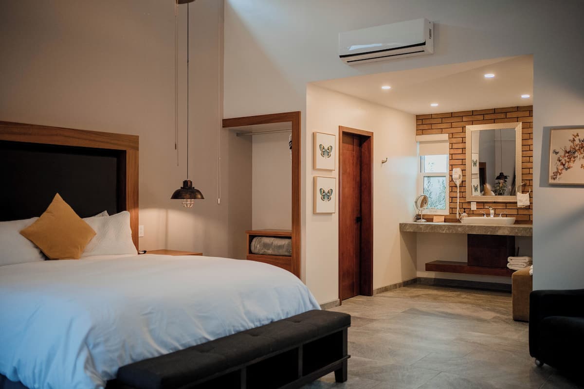 Quarto com cama, um banco, banheiro com pia e espelho, e porta que dá para outro cômodo.