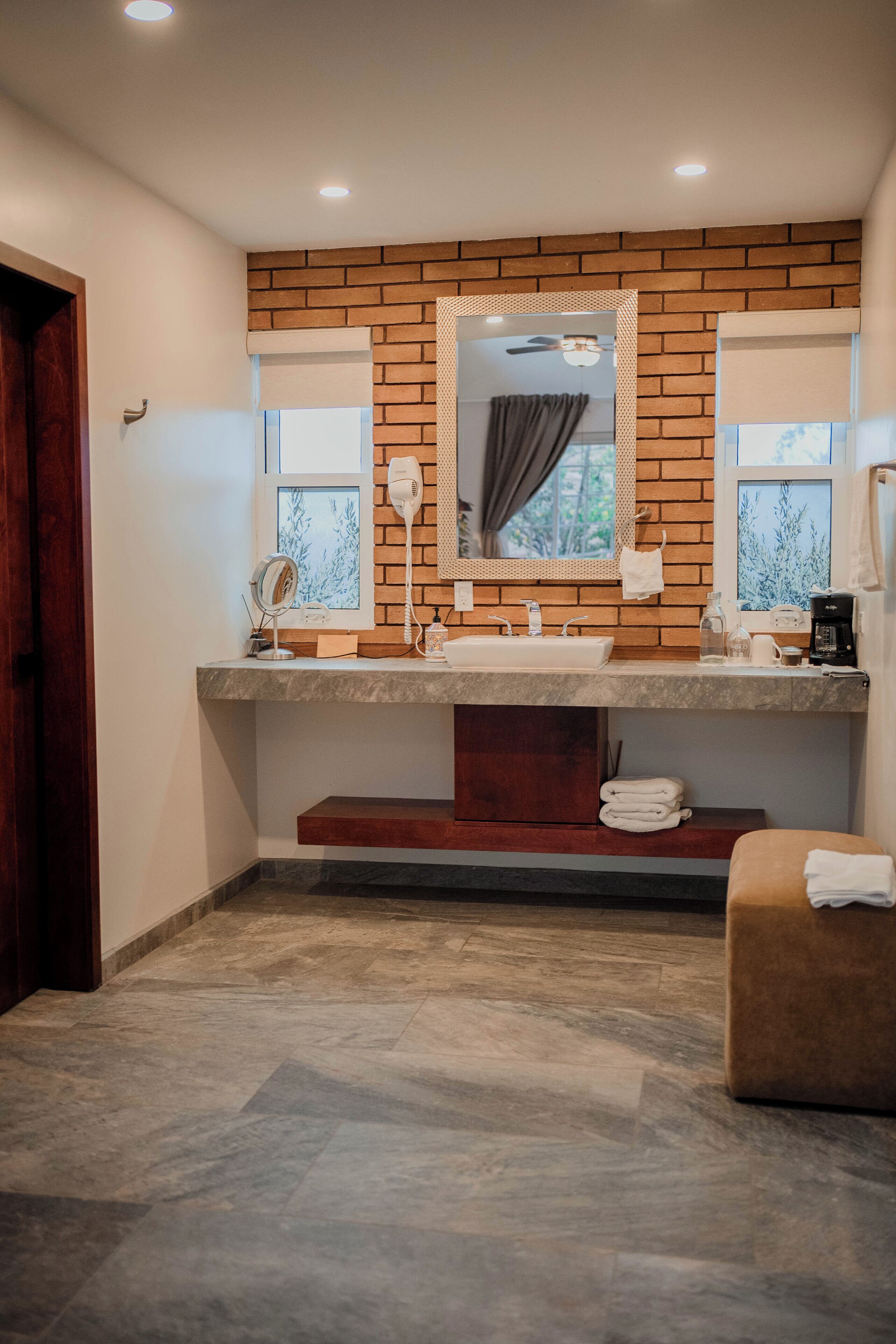 Casa de banho com parede de tijolos, duas pias, um espelho, um porta-toalhas e um banco.