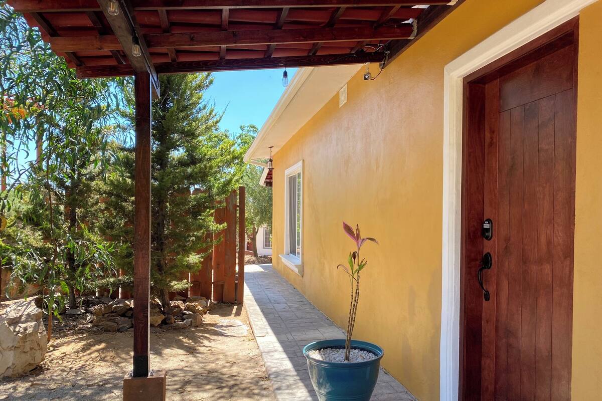 Área externa coberta com pérgola de madeira, planta em vaso e parede amarela com porta de madeira.