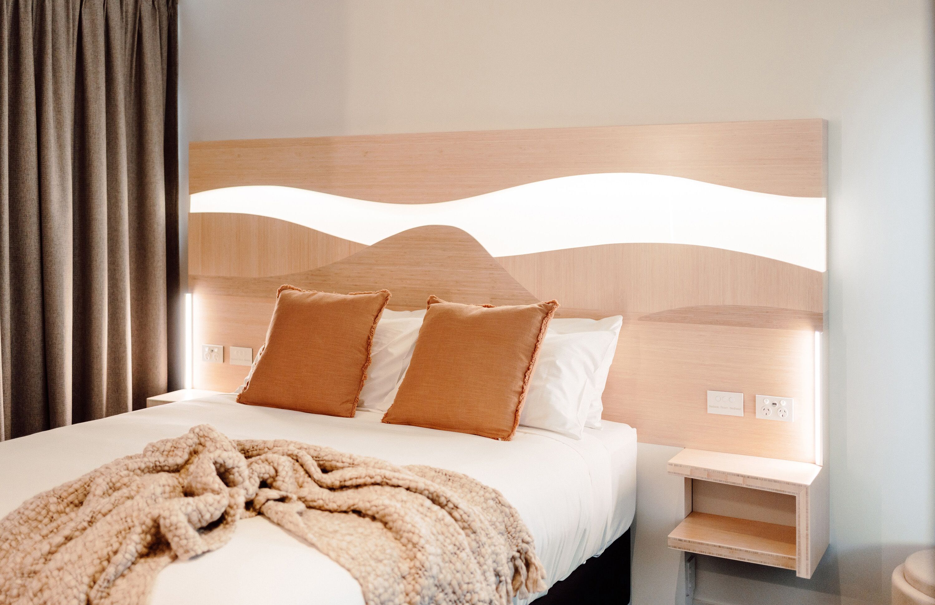 Quarto de hotel moderno com uma cama grande, duas almofadas, uma mesa de cabeceira e um candeeiro fixado na parede.