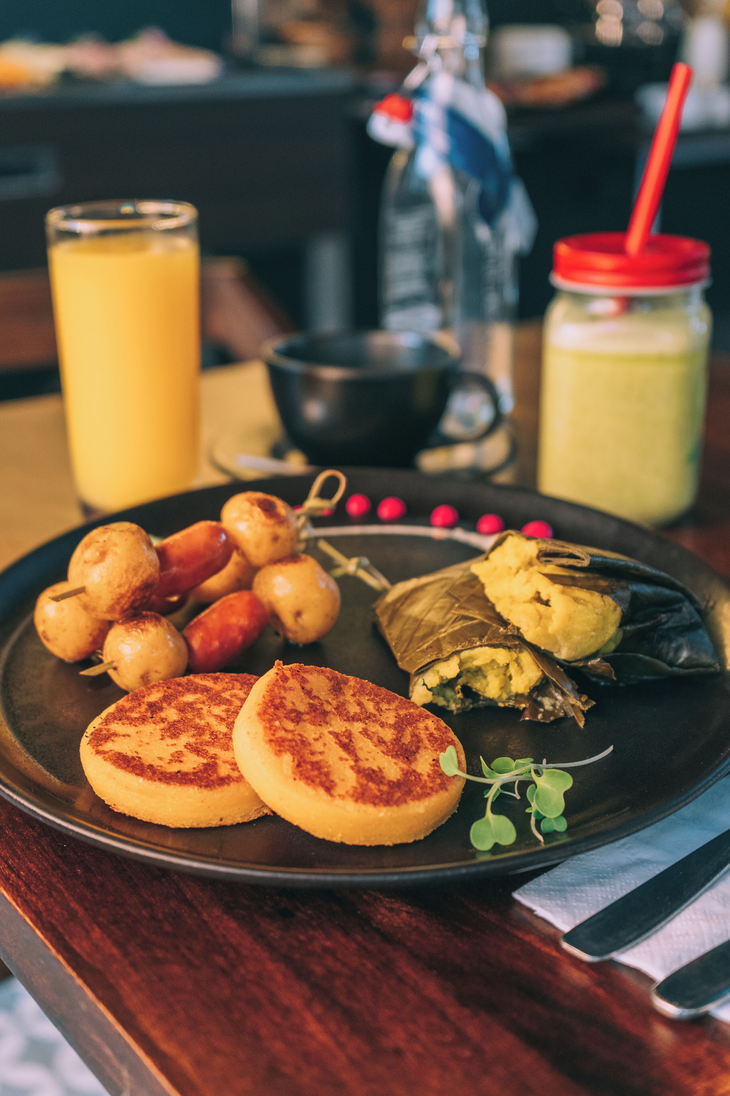 Um prato com alimentos, incluindo salsichas, panquecas e um tamal, acompanhado de um copo de suco de laranja e um pote de molho verde.