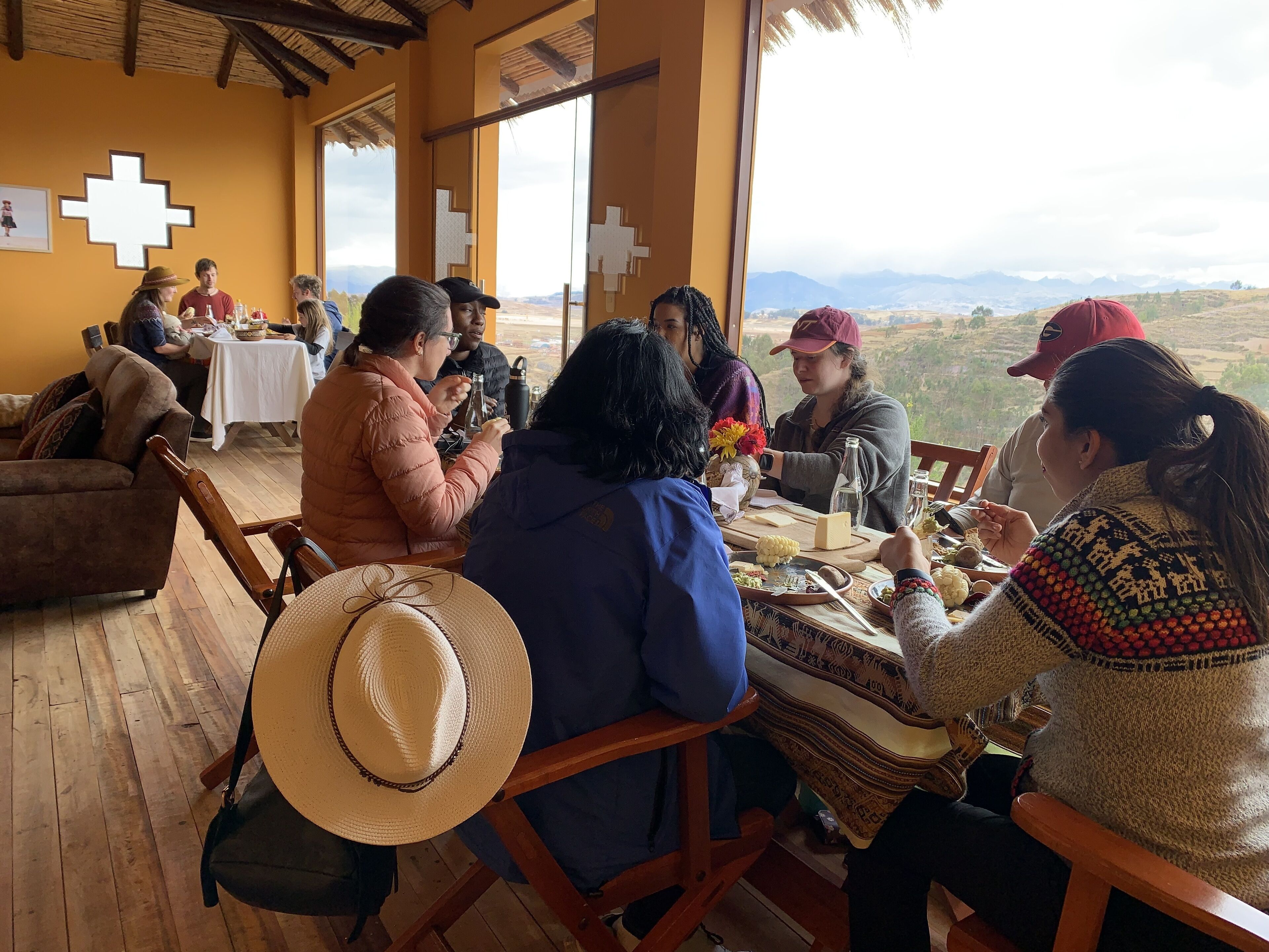 Um grupo de pessoas jantando à mesa com vista para montanhas, móveis de madeira e decoração de parede em formato de cruz.