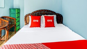 Quarto com cabeceira de madeira, duas almofadas vermelhas com o texto 'OYO', uma cômoda verde e uma cadeira de madeira.