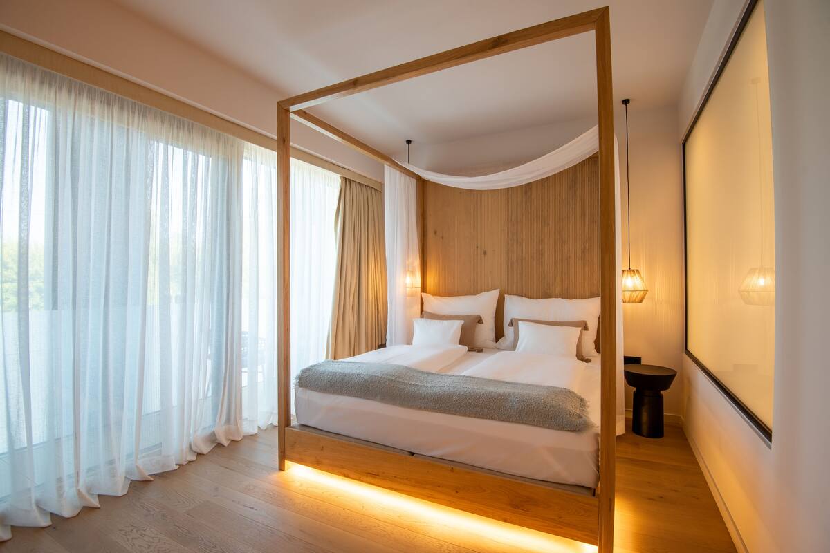 Quarto moderno com estrutura de cama de madeira, roupa de cama branca e cortinas leves.