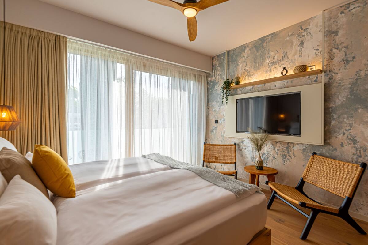 Quarto de hotel moderno com uma cama grande, um ventilador de teto, uma TV na parede e uma janela com cortinas finas.