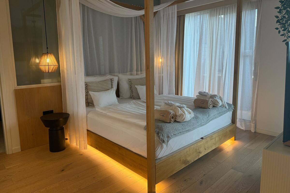 Uma cama de dossel com lençóis brancos, uma mesa de cabeceira e um abajur.
