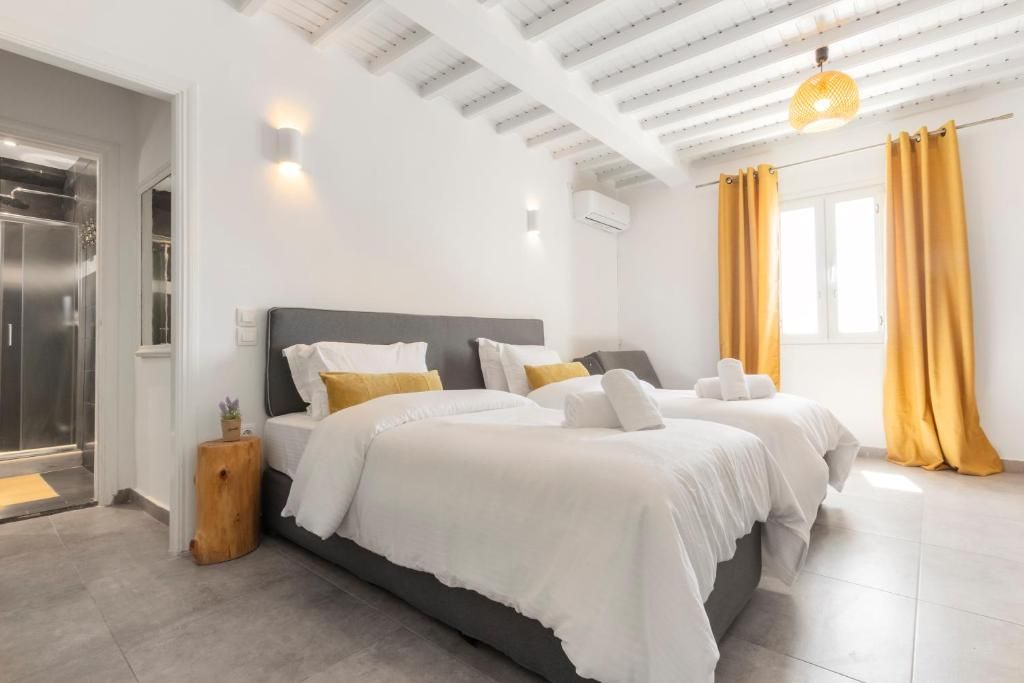 Quarto de hotel com uma cama grande, um banquinho de madeira, uma mesa pequena e uma janela com cortinas.