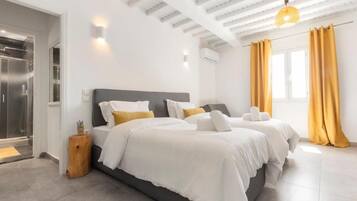 Quarto de hotel com uma cama grande, um banquinho de madeira, uma mesa pequena e uma janela com cortinas.