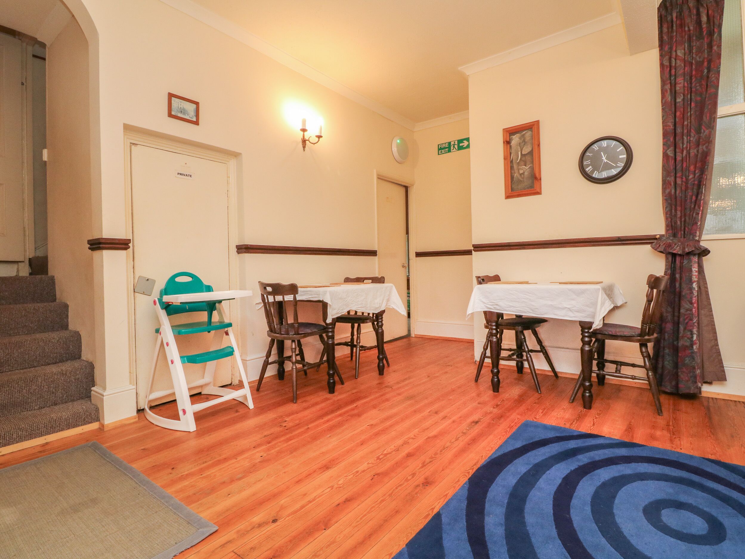 Quarto de hotel com piso de madeira, um tapete azul e preto, uma mesa branca com toalha branca, cadeiras de madeira, uma cadeira verde e branca, uma escada, uma porta, um relógio de parede, uma luminária de parede e o sinal de saída de incêndio.