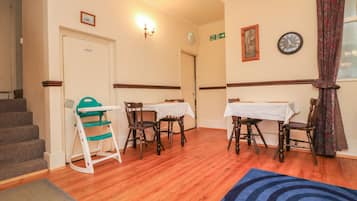 Quarto de hotel com piso de madeira, um tapete azul e preto, uma mesa branca com toalha branca, cadeiras de madeira, uma cadeira verde e branca, uma escada, uma porta, um relógio de parede, uma luminária de parede e o sinal de saída de incêndio.