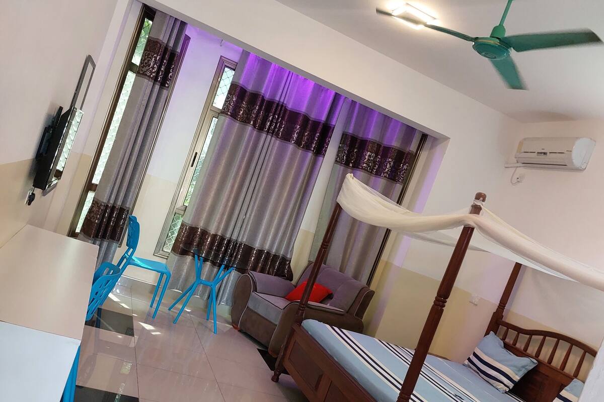 Quarto com cama de dossel, ventilador de teto e janela com iluminação roxa.