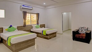 Quarto de hotel com duas camas, um ventilador de teto, aparelho de ar condicionado, janela com cortinas e uma cadeira estampada com flores.