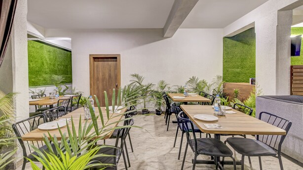 Uma área de refeições interna moderna, com várias mesas de madeira e cadeiras pretas, cercada por plantas verdes e um chão de concreto.