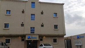 Um edifício de vários andares com fachada bege, entrada central com porta preta e área de estacionamento com um carro branco.