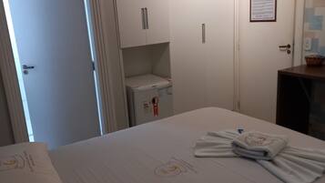 Quarto de hotel com uma cama, guarda-roupa, uma pequena mesa e uma prateleira fixada na parede.