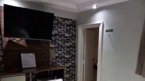 Quarto de hotel com televisão de tela plana fixada em parede com padrão de tijolos, minibar, porta identificada com "Cabideiro" e camas bem arrumadas com toalhas.