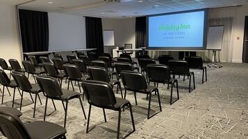 Uma sala de conferências com fileiras de cadeiras, uma tela de projetor exibindo "Holiday Inn" e uma lousa.