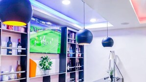 Um espaço de bar moderno com uma televisão de tela plana exibindo uma partida de futebol, prateleiras com garrafas e luminárias pendentes.