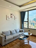 Uma sala de estar moderna com um sofá cinzento, uma mesinha redonda com um vaso de flores e vista para a cidade através de amplas janelas.