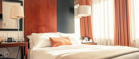 Quarto de hotel com cama, mesas de cabeceira, abajur e cortinas.