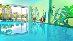 Piscina interior com água cristalina, rodeada por paredes decoradas com plantas tropicais e dois boias salva-vidas vermelhas.