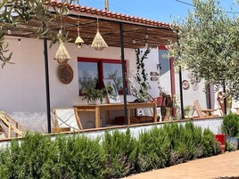 Um edifício com telhado de telhas vermelhas, varanda coberta com mobiliário de madeira e um jardim com diversas plantas.
