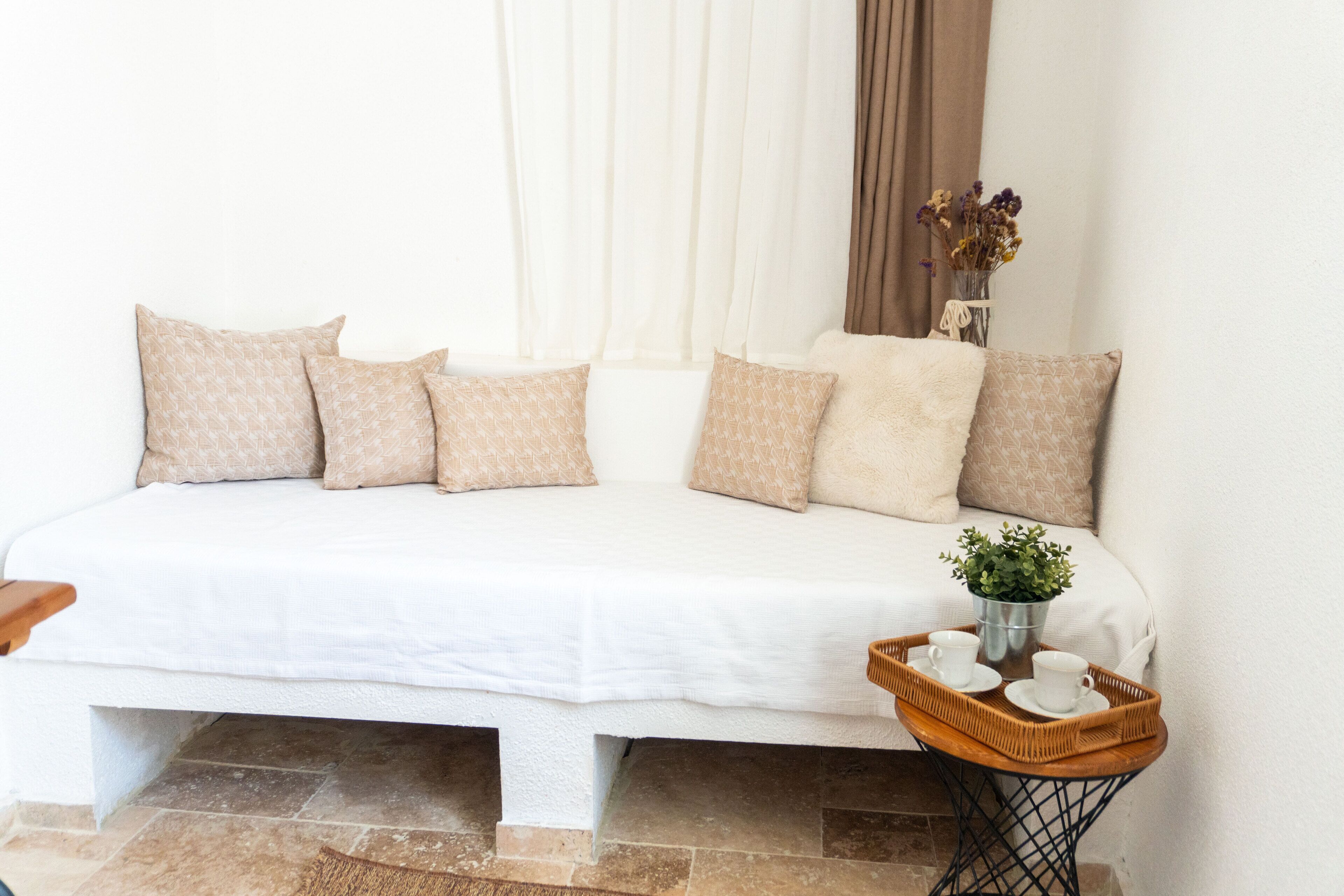 Cama arrumada com almofadas estampadas, mesa de cabeceira pequena com bandeja de xícaras e um vaso de plantas, e um vaso de flores secas em uma parede branca.