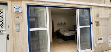 Entrada de hotel com porta de vidro, cama com colcha estampada e uma pequena prateleira com objetos decorativos.