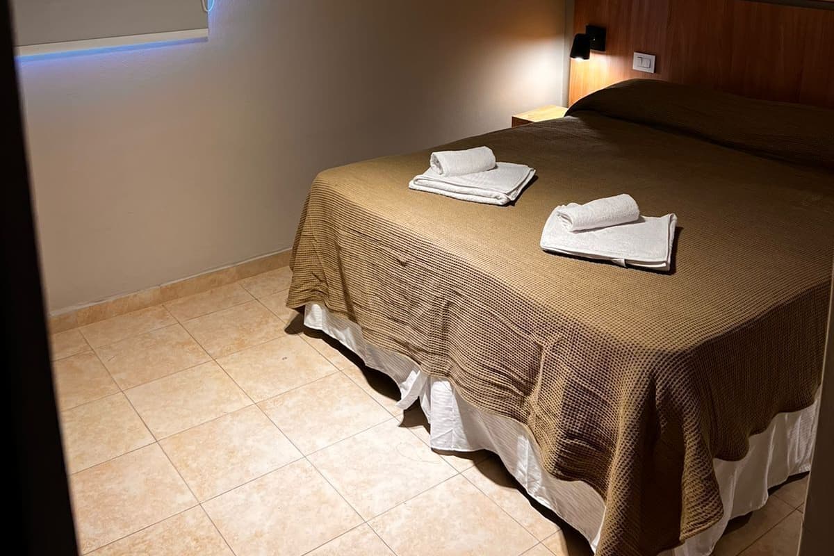 Quarto de hotel com uma cama, uma janela, um quadro na parede e um candeeiro no teto.